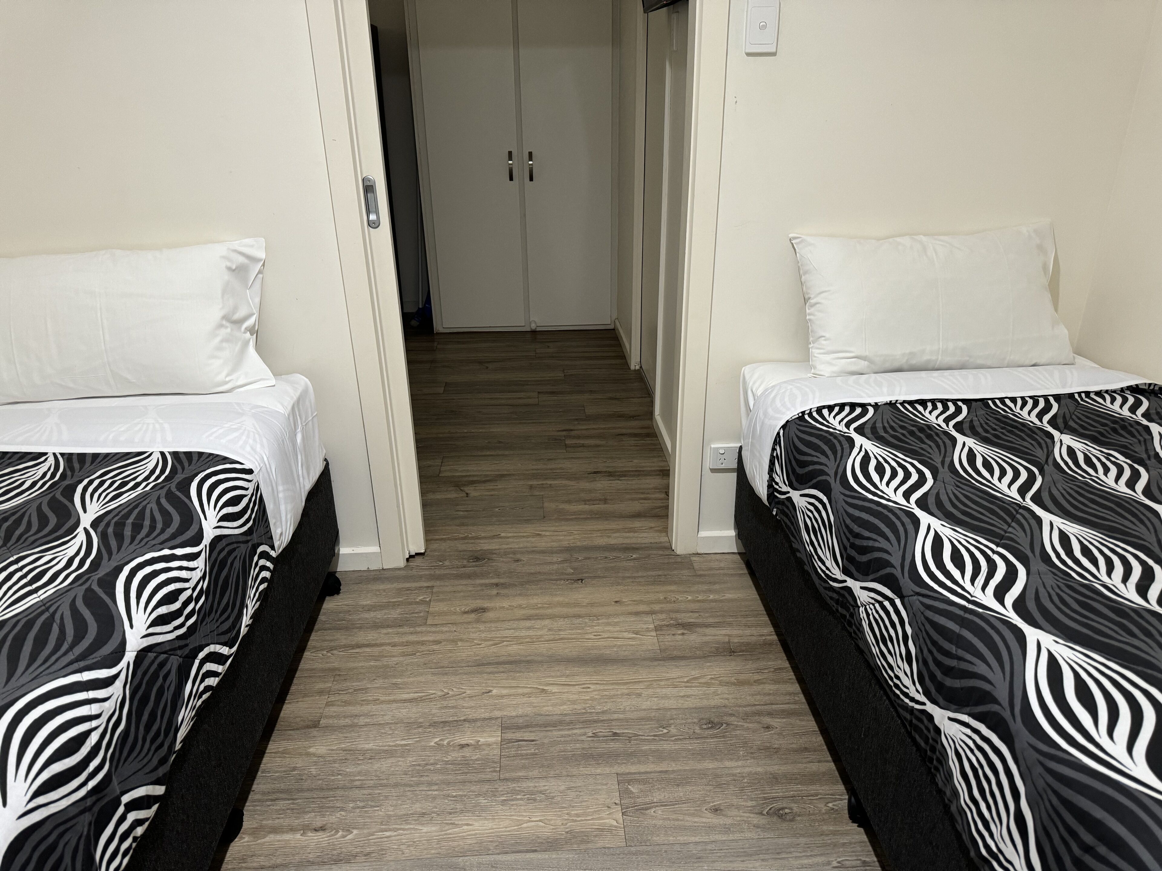 Um corredor que dá para um quarto com duas camas, cada uma com colcha estampada e almofadas brancas.
