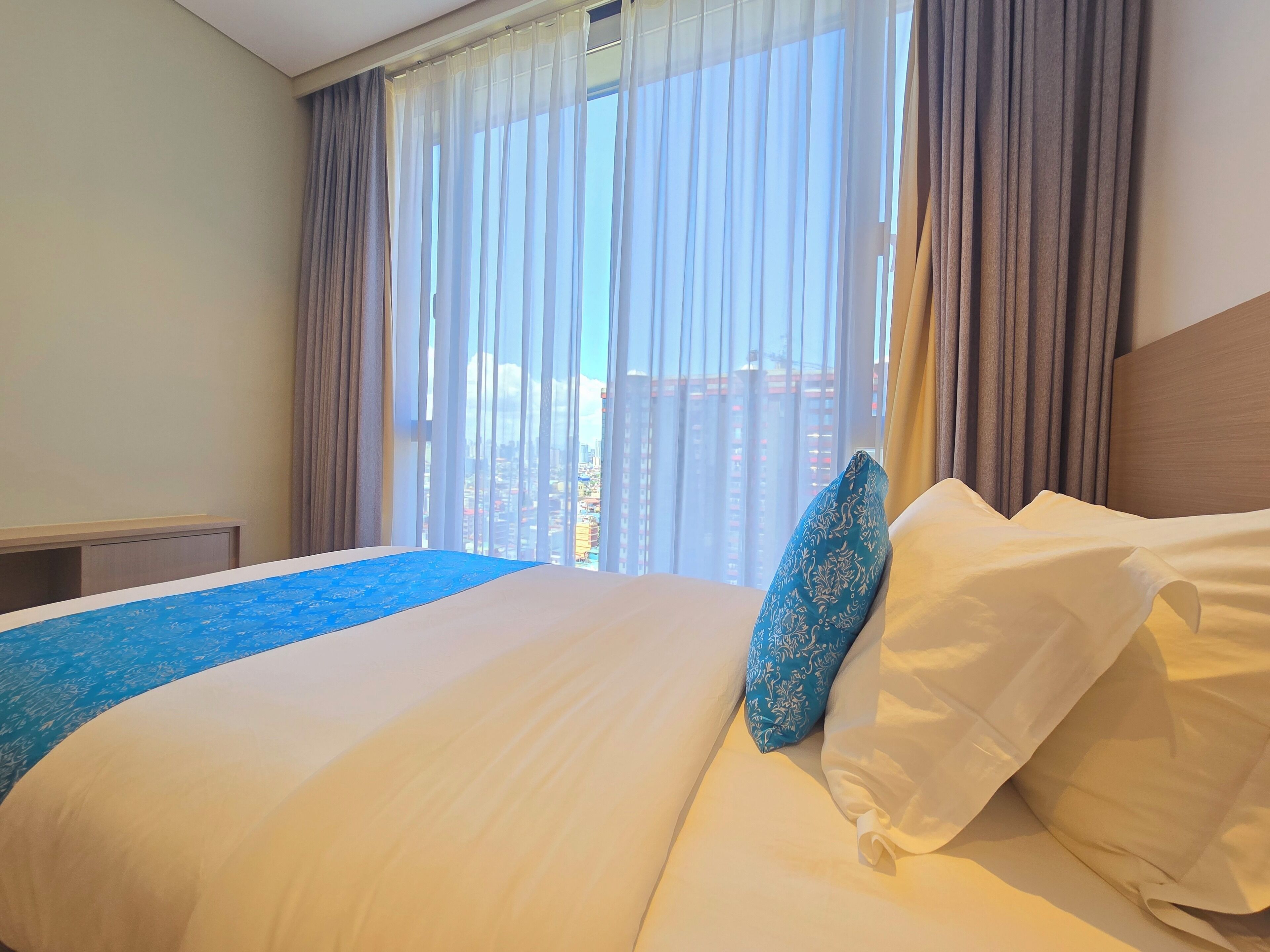 Quarto de hotel com cama, vista da cidade através de cortinas translúcidas e cabeceira de madeira.