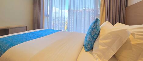 Quarto de hotel com cama, vista da cidade através de cortinas translúcidas e cabeceira de madeira.