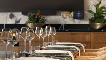 Mesa de jantar posta com taças de vinho, pratos e talheres, com uma televisão e plantas decorativas ao fundo.