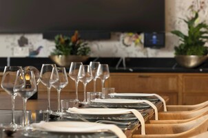 Mesa de jantar posta com taças de vinho, pratos e talheres, com uma televisão e plantas decorativas ao fundo.