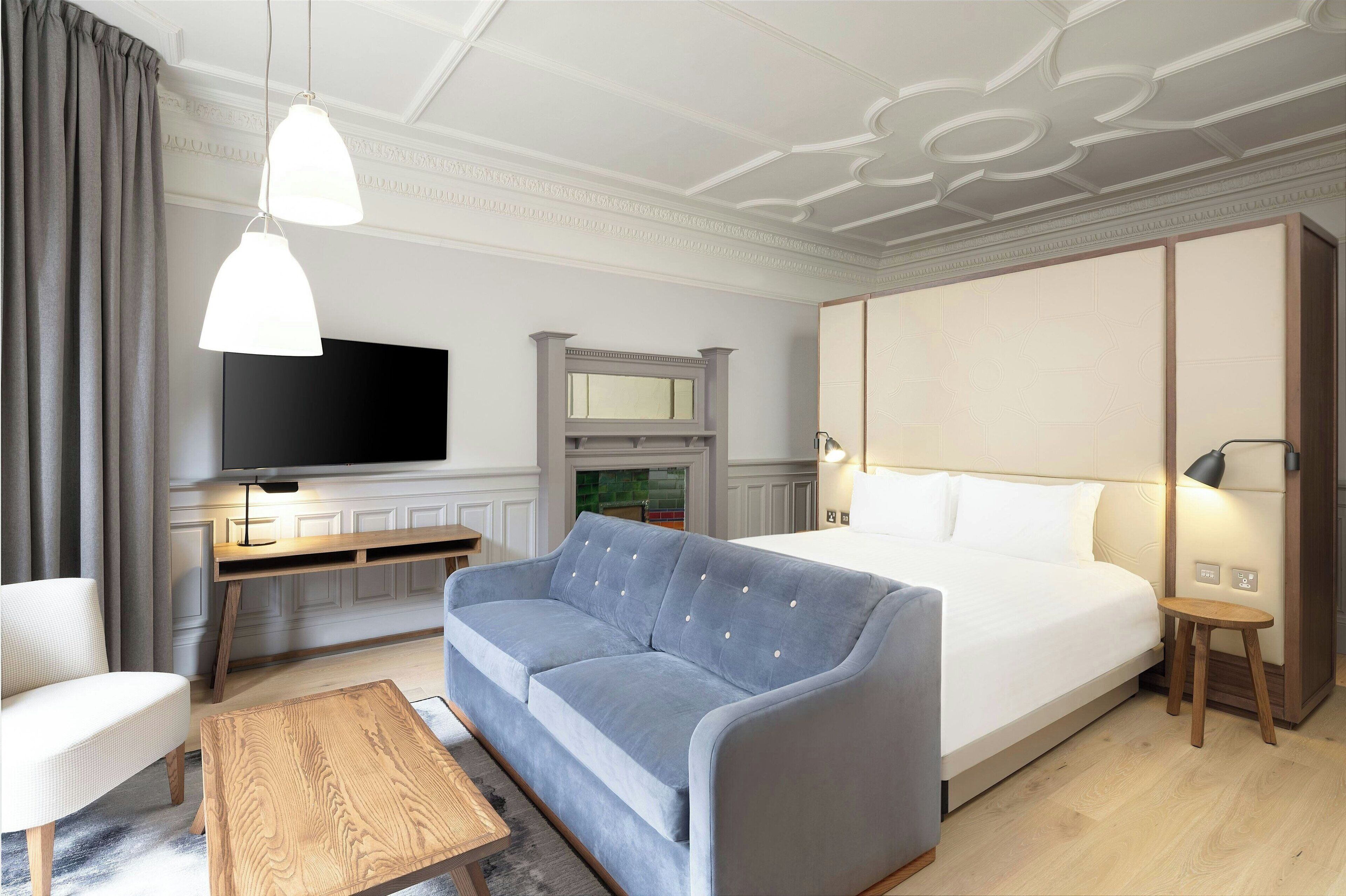 Quarto de hotel moderno com sofá cinzento, mesa de centro de madeira, cama, televisão e pequena mesa de cabeceira com abajur.