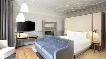 Quarto de hotel moderno com sofá cinzento, mesa de centro de madeira, cama, televisão e pequena mesa de cabeceira com abajur.