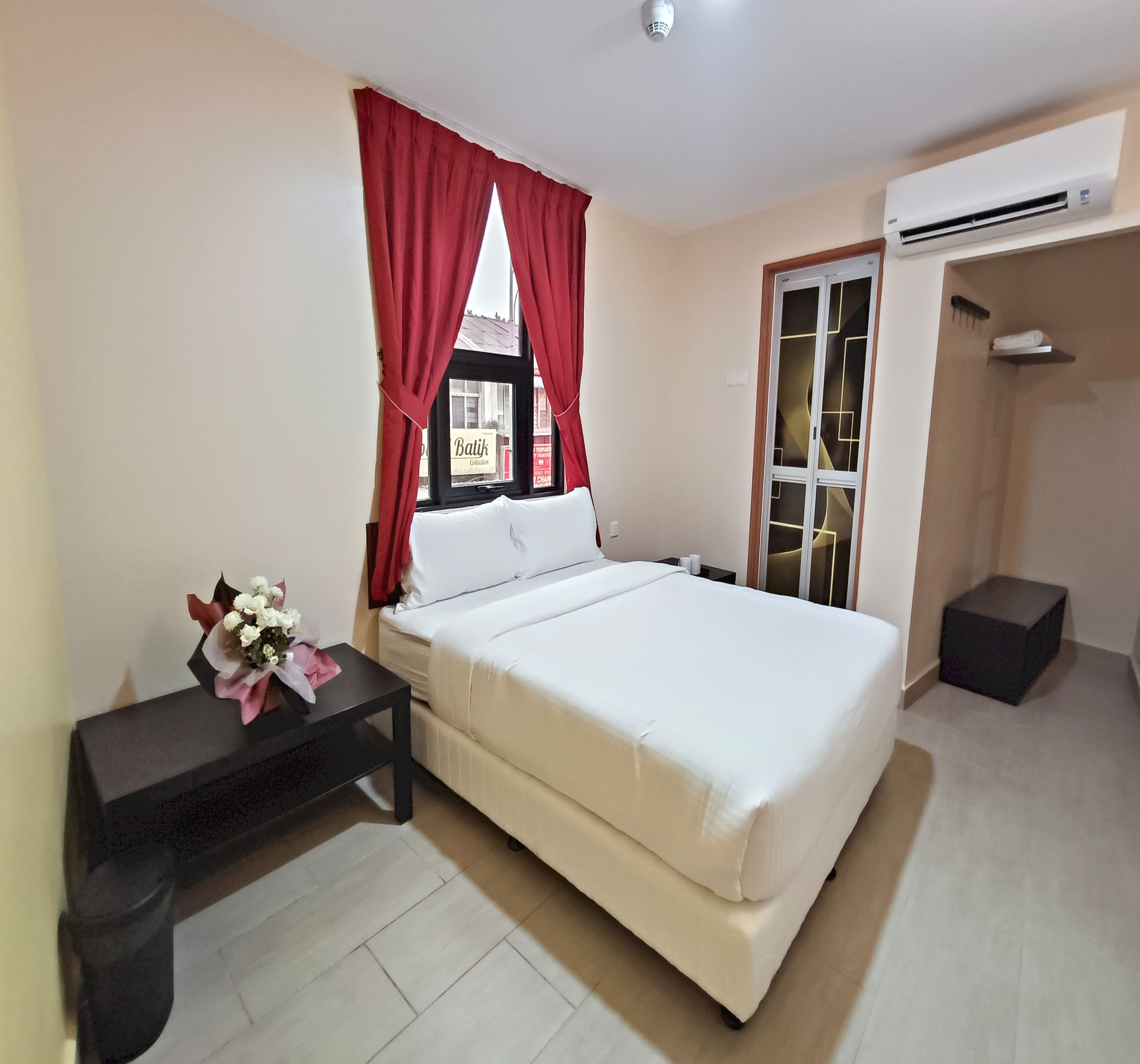 Quarto com cama, mesa de cabeceira, janela com cortinas vermelhas, aparelho de ar condicionado e um pequeno banquinho.