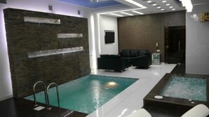 Uma área moderna de piscina interna com parede de pedra, um espaço de estar com sofás pretos e uma banheira de hidromassagem separada.
