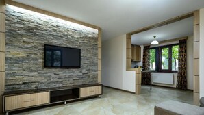Uma sala de estar moderna com uma parede de pedra, uma TV de tela plana, um móvel embutido para entretenimento e uma área de cozinha com janela e cortinas.