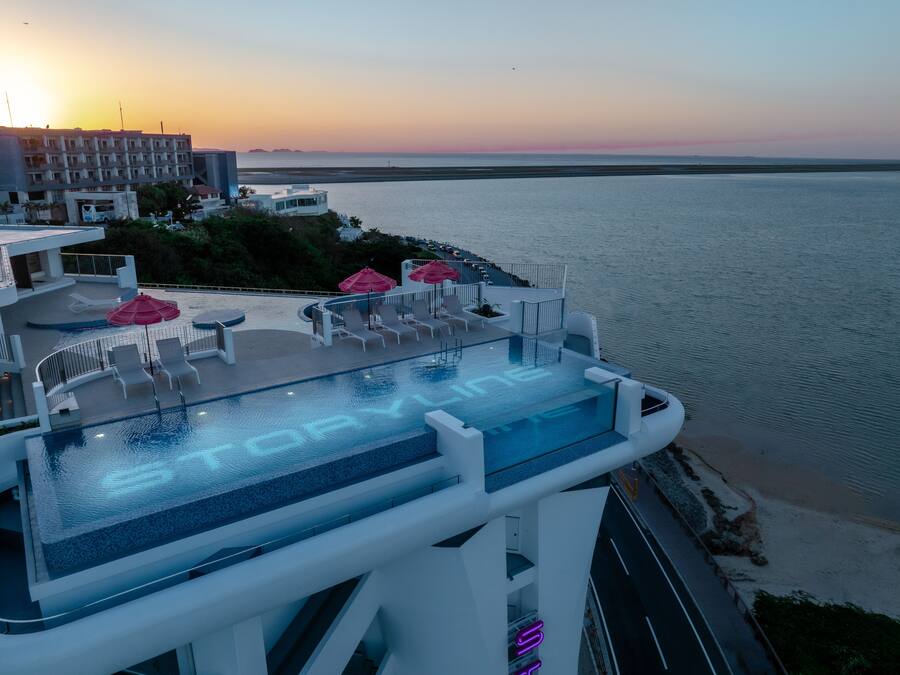 Piscina infinita no terraço com espreguiçadeiras e guarda-sóis, com vista para uma praia e oceano ao pôr do sol.