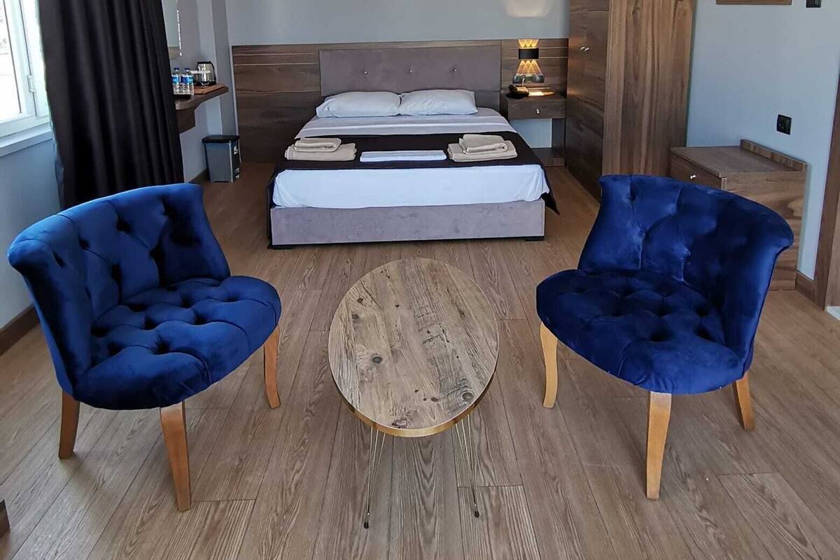 Quarto de hotel moderno com uma cama, duas poltronas azuis, uma mesa de centro redonda e uma TV na parede.