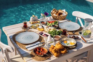 Mesa posta com itens de café da manhã, incluindo pão, ovos, azeitonas e vegetais, à beira de uma piscina.