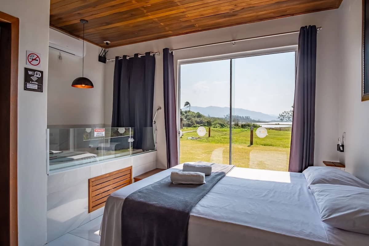 Quarto com uma janela grande, uma cama, teto de madeira e vista para uma paisagem verde.