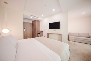 Quarto de hotel moderno com uma cama grande, uma TV de tela plana na parede, uma escrivaninha pequena e um sofá.