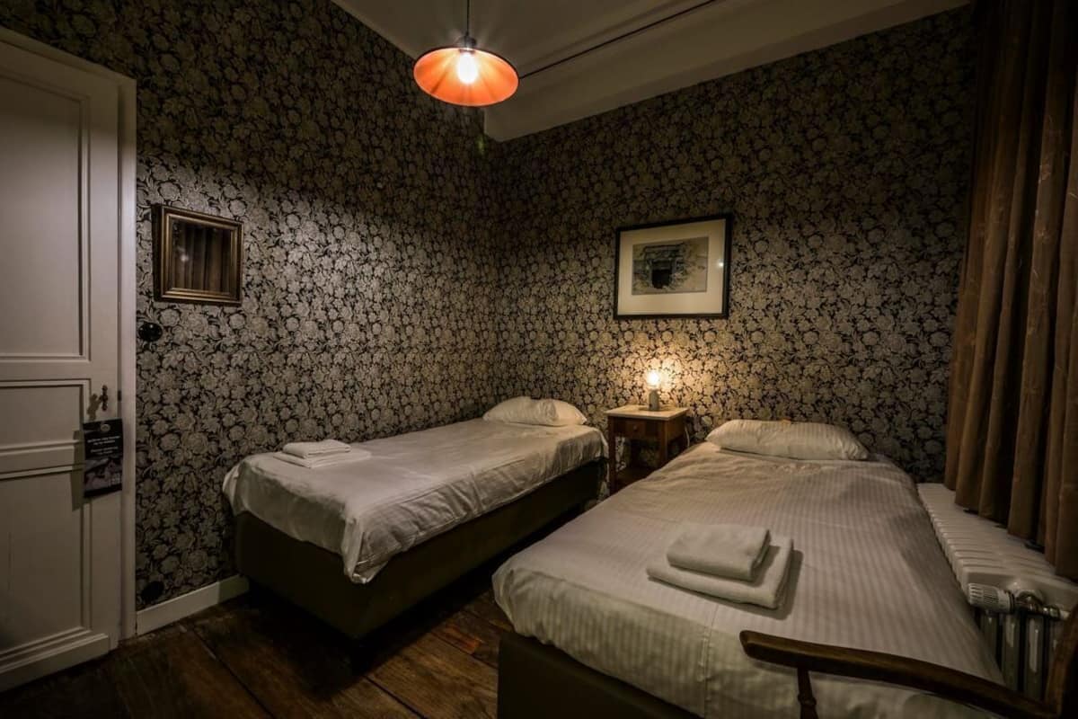 Quarto com duas camas de solteiro, uma mesinha de cabeceira com abajur e uma porta com placa.