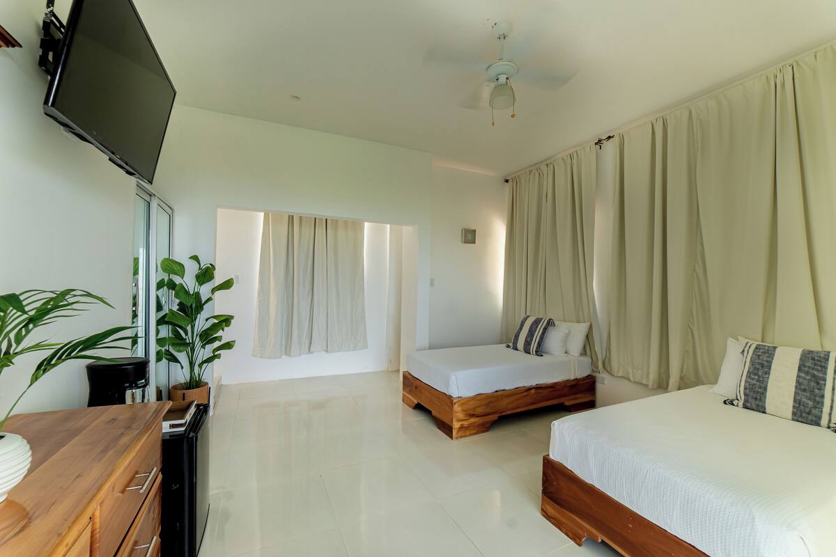 Quarto de hotel com cama, televisão, cômoda de madeira, planta e janela com cortinas.