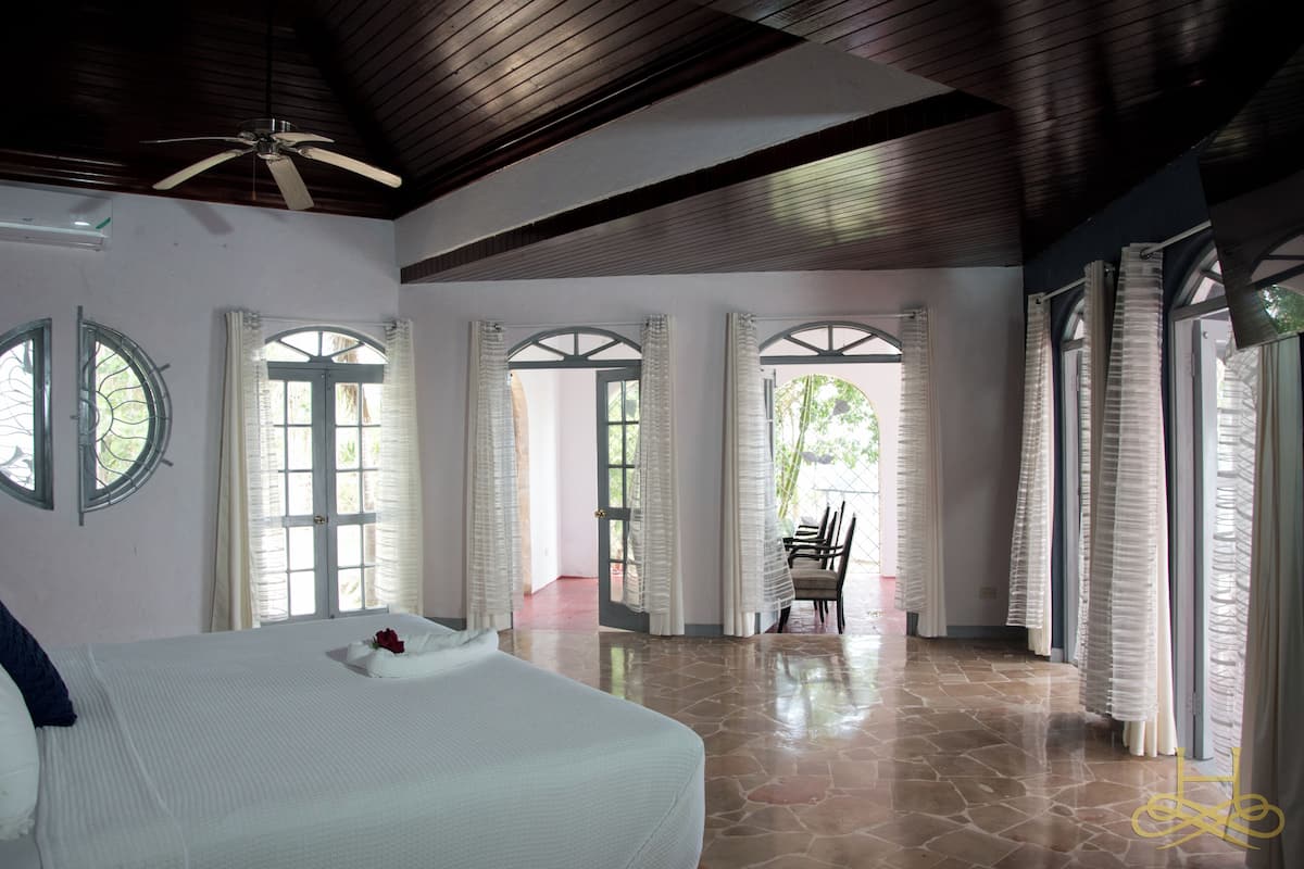 Um quarto amplo com uma cama grande, um ventilador de teto e várias janelas com cortinas.