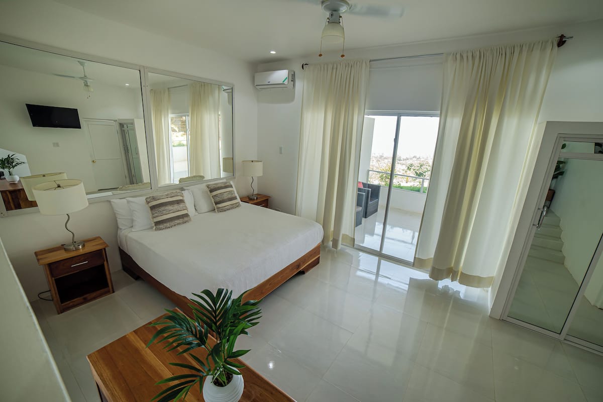 Quarto com uma cama grande, mesa de cabeceira com abajur, espelho e porta de vidro deslizante para uma varanda.