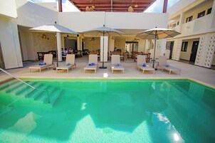 Uma área à beira da piscina com espreguiçadeiras e guarda-sóis, uma piscina cristalina e um espaço coberto com mesas e cadeiras.