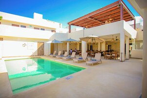 Uma área de piscina com espreguiçadeiras e guarda-sóis, um espaço de estar coberto e um edifício com vários andares e janelas.
