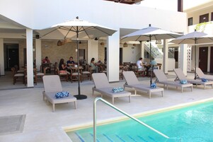 Área da piscina com espreguiçadeiras e guarda-sóis, uma piscina e um espaço de refeições com pessoas.