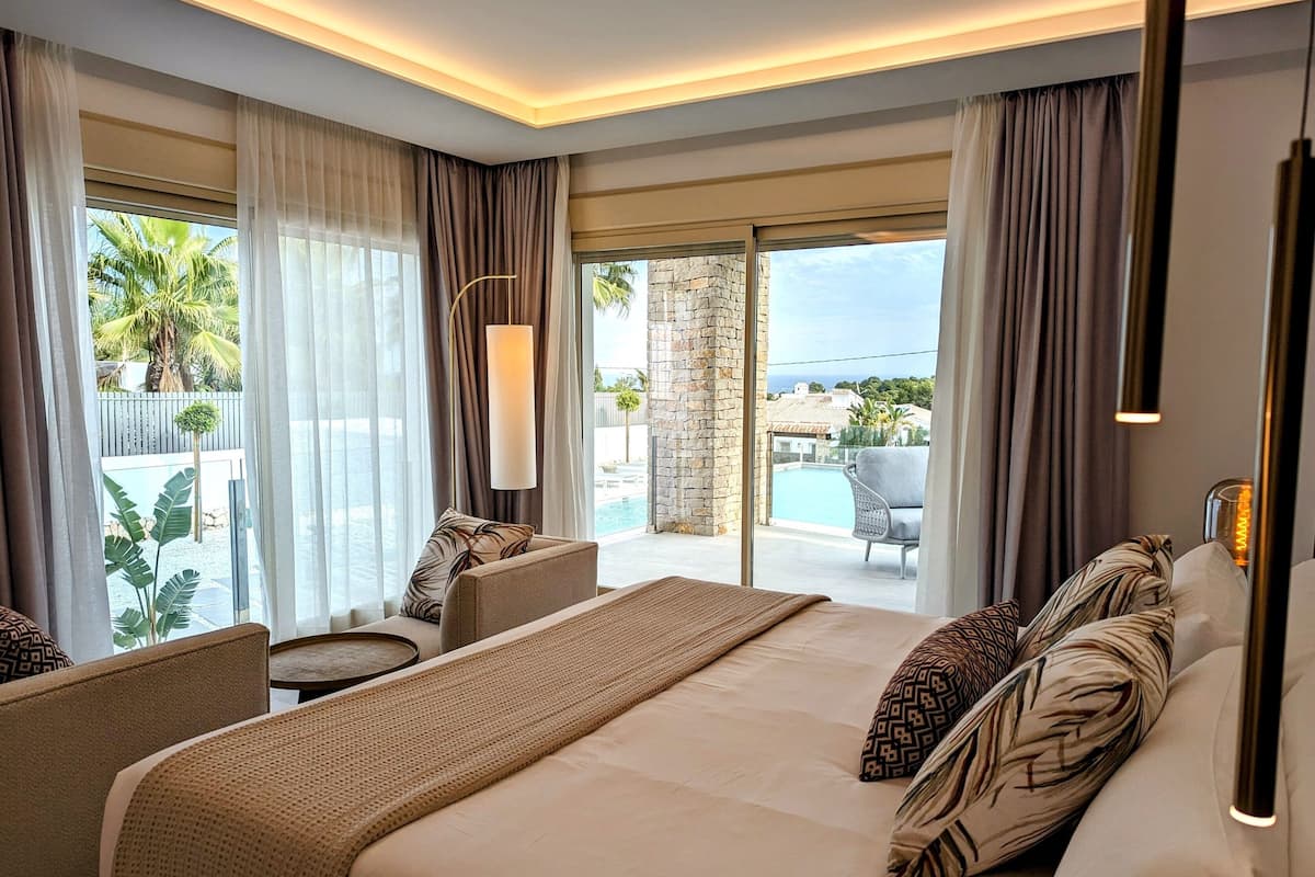 Quarto de hotel com uma cama grande, duas poltronas, uma mesinha e vista para uma piscina e palmeiras.