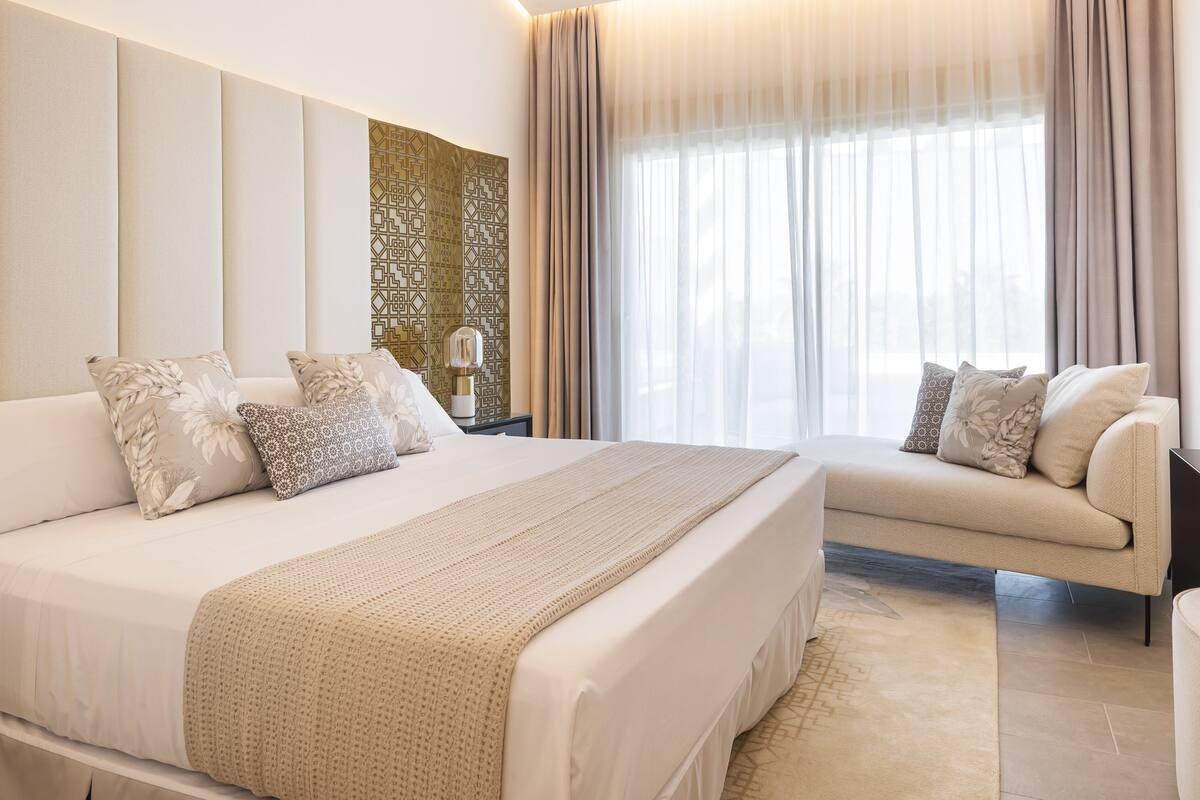 Quarto de hotel moderno com uma cama grande, um sofá, uma mesa de cabeceira com abajur e uma janela ampla com cortinas.