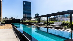 Piscina no terraço com espreguiçadeiras e um pergolado, cercada por edifícios da cidade.