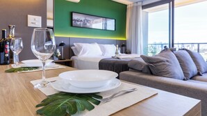 Quarto de hotel com mesa de jantar posta para dois, cama com roupa de cama cinza, parede de destaque verde e uma grande janela com vista para a cidade.