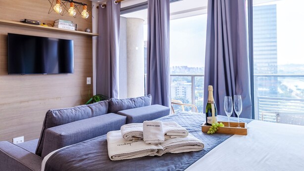 Quarto de hotel moderno com uma cama grande, televisão de tela plana, um sofá e vista para a cidade.