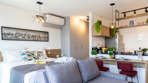 Quarto de hotel moderno com cama, sofá cinza, área de refeições com cozinha e uma obra de arte com vista para a cidade na parede.