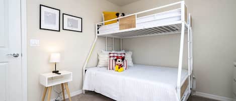 Uma cama beliche com um travesseiro do Mickey Mouse, uma mesinha de cabeceira branca e pequena com uma luminária, e duas fotos emolduradas na parede.