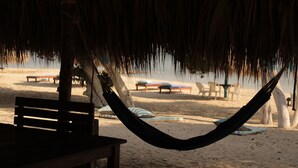 Uma rede sob um telhado de palha, um banco de madeira e cadeiras de praia com almofadas listradas numa praia de areia.