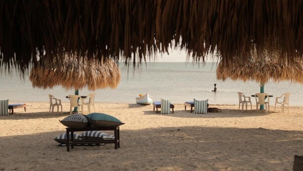 Cenário de praia com guarda-sóis de palha, espreguiçadeiras e uma mesa com almofadas.