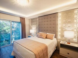 Quarto com cama grande, duas mesas de cabeceira com luminárias, cabeceira decorativa e vista para o verde através de uma porta de correr.