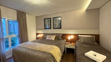 Quarto de hotel com duas camas, uma janela grande com cortinas, um candelabro e duas obras de arte emolduradas na parede.