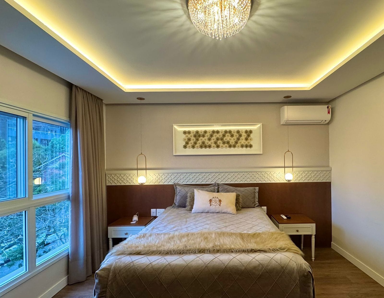 Quarto moderno com uma cama grande, duas mesas de cabeceira, um candelabro, ar condicionado e uma janela com cortinas.