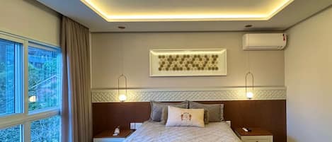 Quarto moderno com uma cama grande, duas mesas de cabeceira, um candelabro, ar condicionado e uma janela com cortinas.