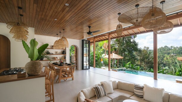 Uma sala de estar moderna com teto de madeira, luminárias de fibra natural, uma planta grande, um conjunto de jantar e vista para uma piscina.