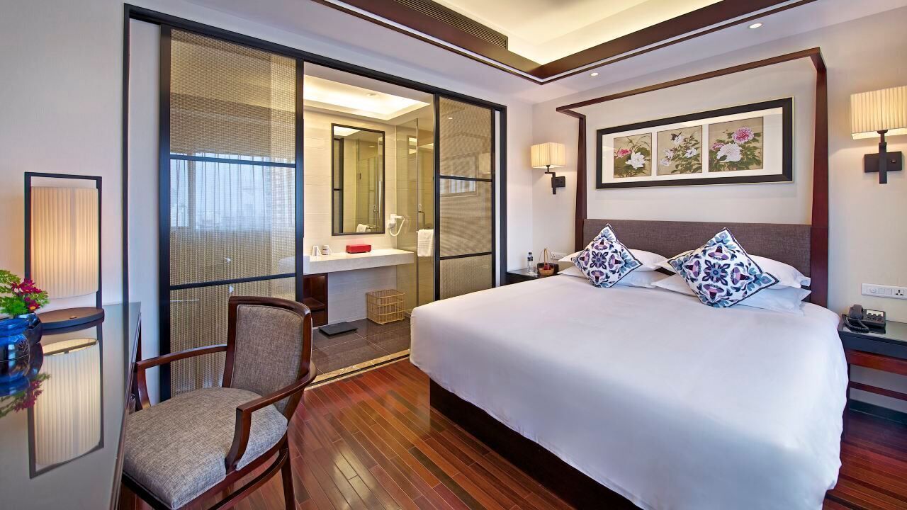 Quarto de hotel com uma cama grande, uma cadeira, uma escrivaninha, um espelho e uma porta de correr.