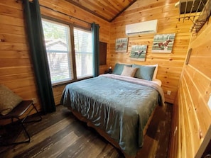 Quarto de cabana de madeira com cama, um banco e janela com cortinas.