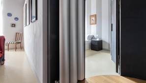 Um corredor com uma cadeira de madeira, uma cama com travesseiro branco e uma mesa de cabeceira com abajur.