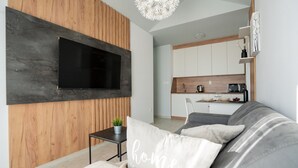 Sala de estar moderna com sofá cinza, televisão de tela plana fixada numa parede de madeira, uma mesinha com uma planta e uma área de cozinha ao fundo.
