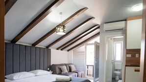Quarto com uma cama grande, vigas de madeira no teto, um banheiro pequeno e vista para o exterior.