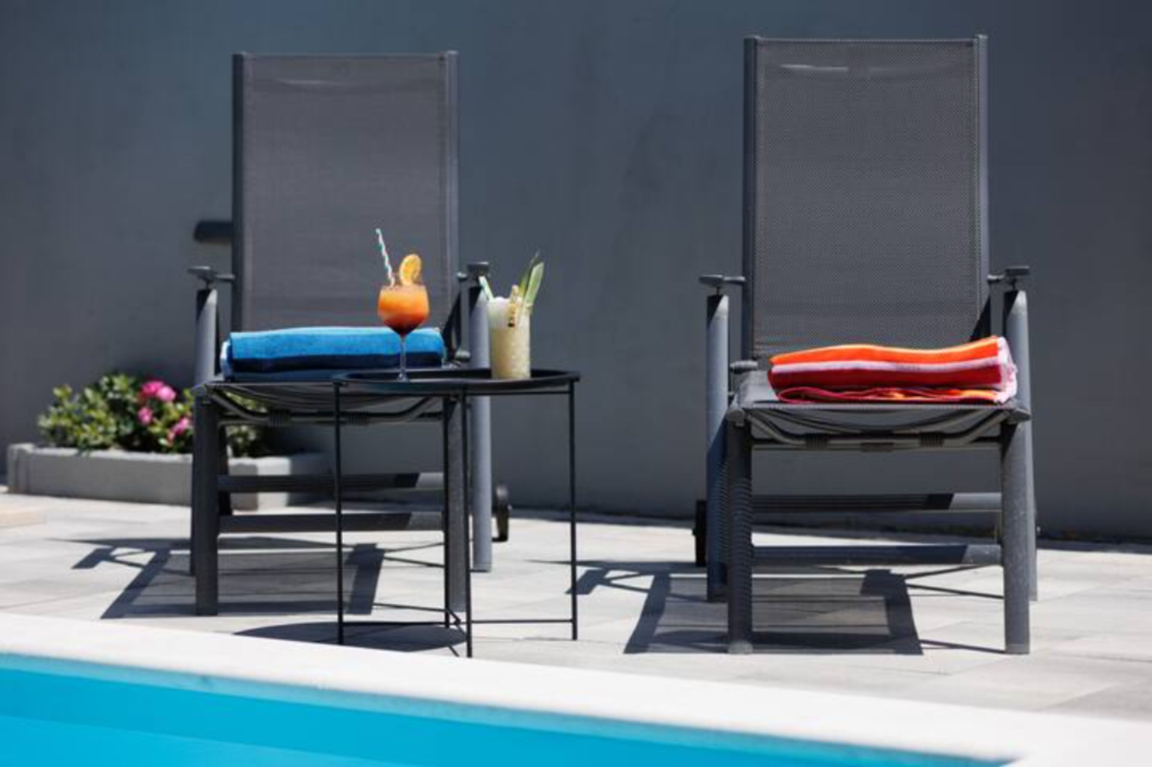 Duas cadeiras de descanso ao ar livre com almofadas, uma mesa pequena com bebida e uma vela, e uma piscina ao fundo.