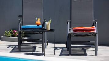 Duas cadeiras de descanso ao ar livre com almofadas, uma mesa pequena com bebida e uma vela, e uma piscina ao fundo.