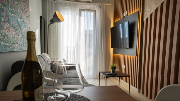 Uma sala de estar moderna com um sofá, uma mesa de jantar com garrafa e copo, uma TV fixada em uma parede de madeira e uma janela com cortinas leves.