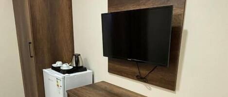 Quarto compacto com guarda-roupa de madeira, eletrodoméstico pequeno branco, escrivaninha de madeira e televisão de tela plana fixada na parede.