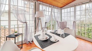 Uma sala de jantar com uma mesa redonda, cadeiras, uma luminária e vista para o verde através de grandes janelas.