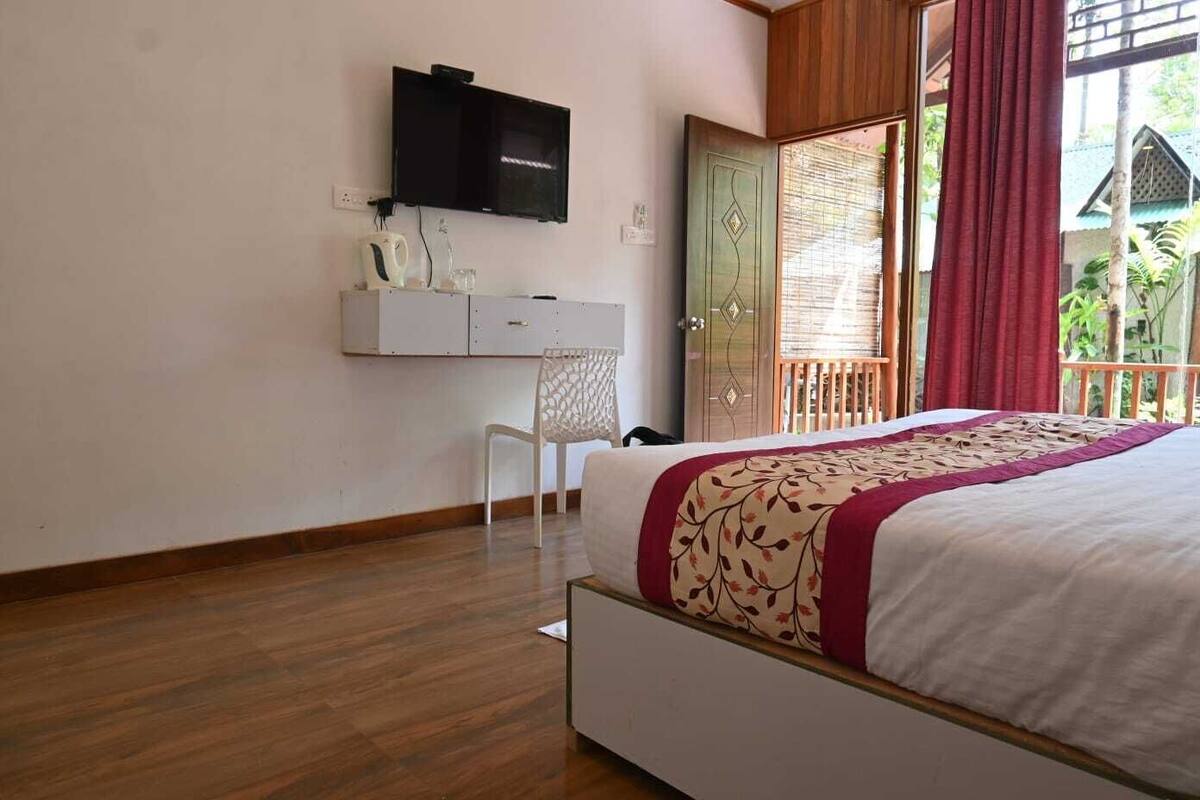 Quarto com cama, televisão na parede, escrivaninha e janela com vista para vegetação.