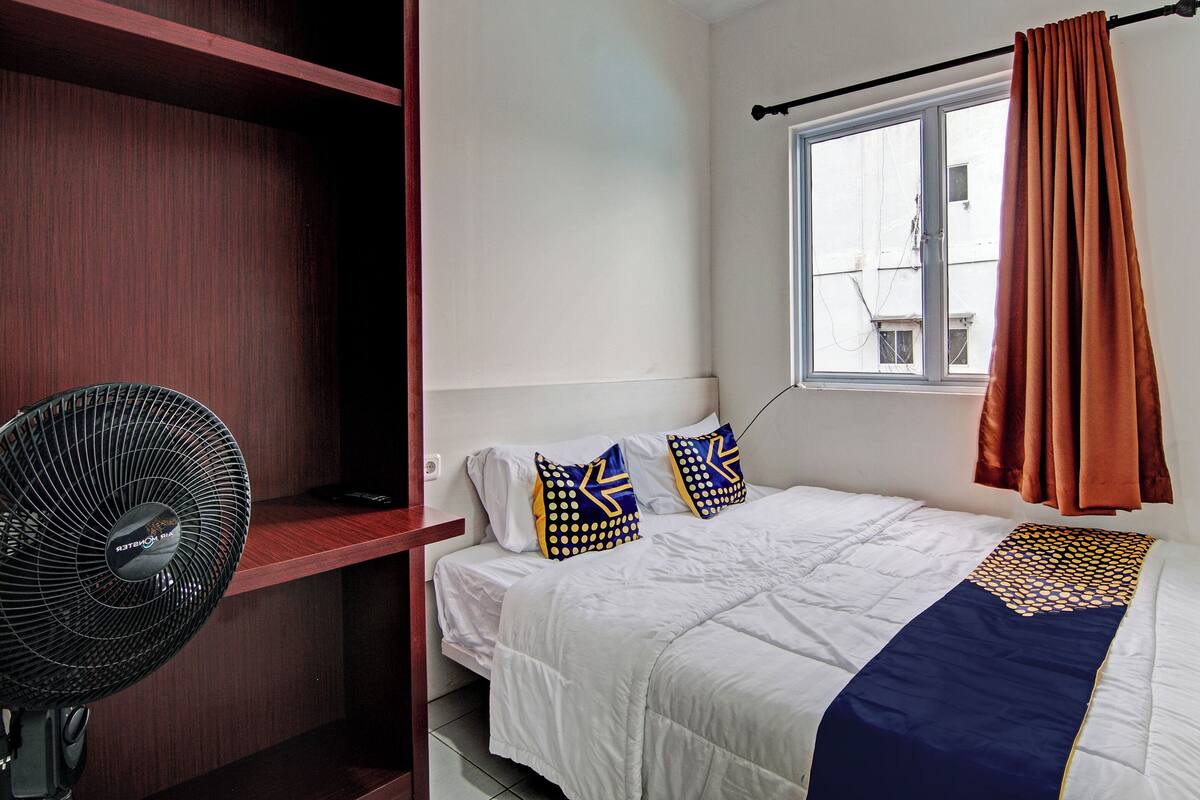 Quarto com cama, um ventilador, janela com cortinas e prateleira de madeira.