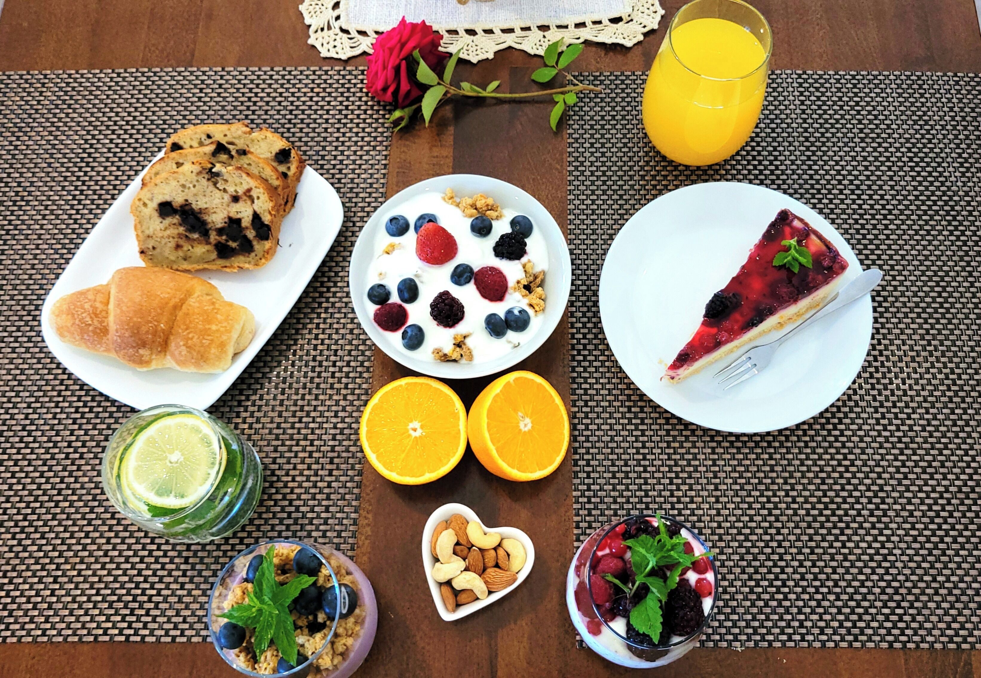 Mesa de café da manhã com croissants, uma fatia de cheesecake, iogurte com frutas vermelhas, uma tigela em formato de coração com amêndoas, uma tigela com frutas vermelhas sortidas e um copo de suco de laranja.