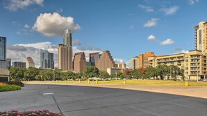 Paisagem urbana com edifícios altos e modernos, uma ampla praça e uma área de parque com árvores e flores.